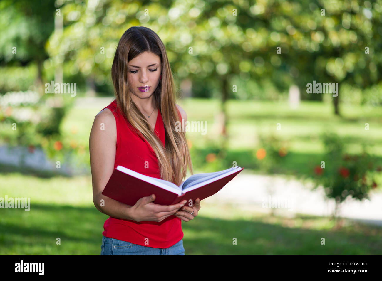 Portrait of a young blonde girl reading a red book in a park in spring ...