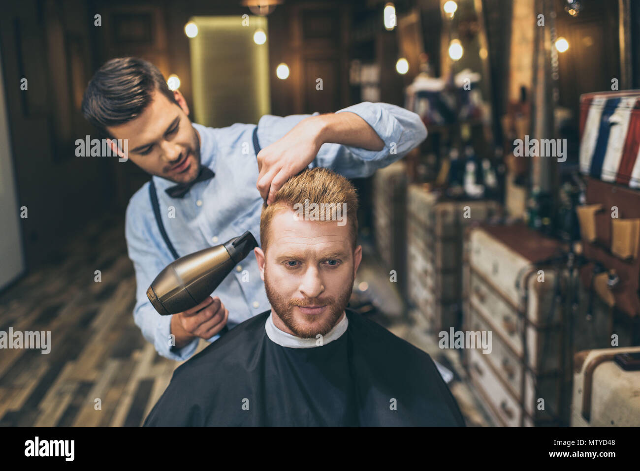 Young barber drying hair of customer with hair dryer in barber shop ...