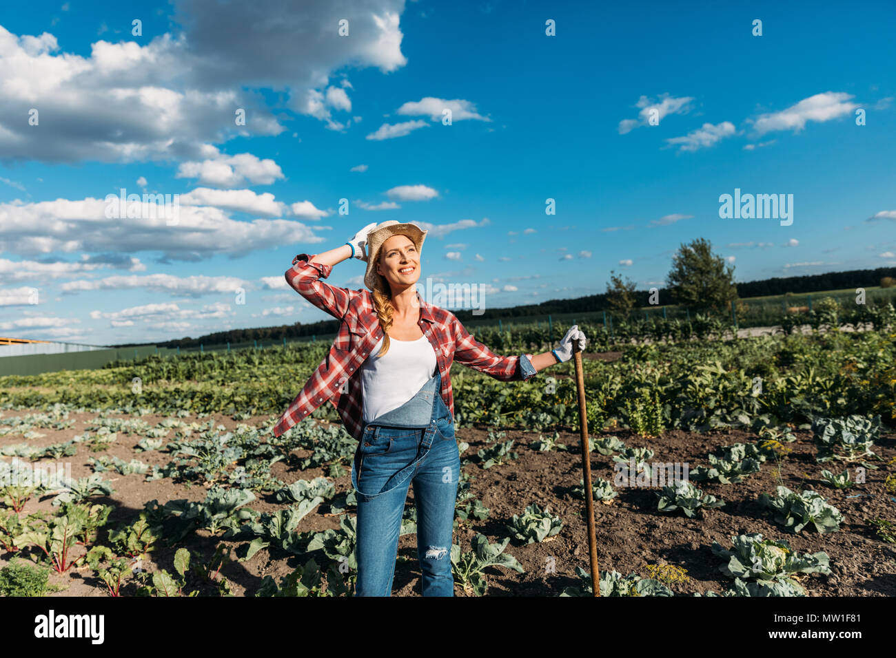 beautiful young female farmer in hat holding hoe and working on field ...
