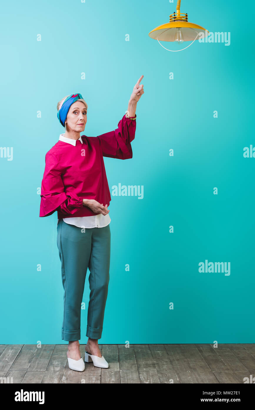 fashionable senior woman pointing at lamp in room with turquoise wall ...