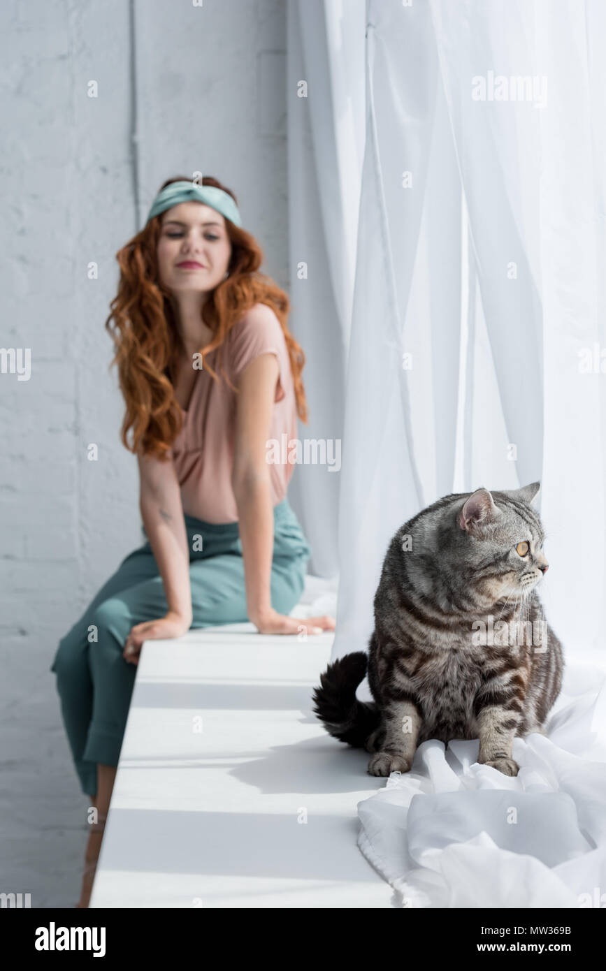 happy young woman sitting on windowsill with tabby cat on foreground ...