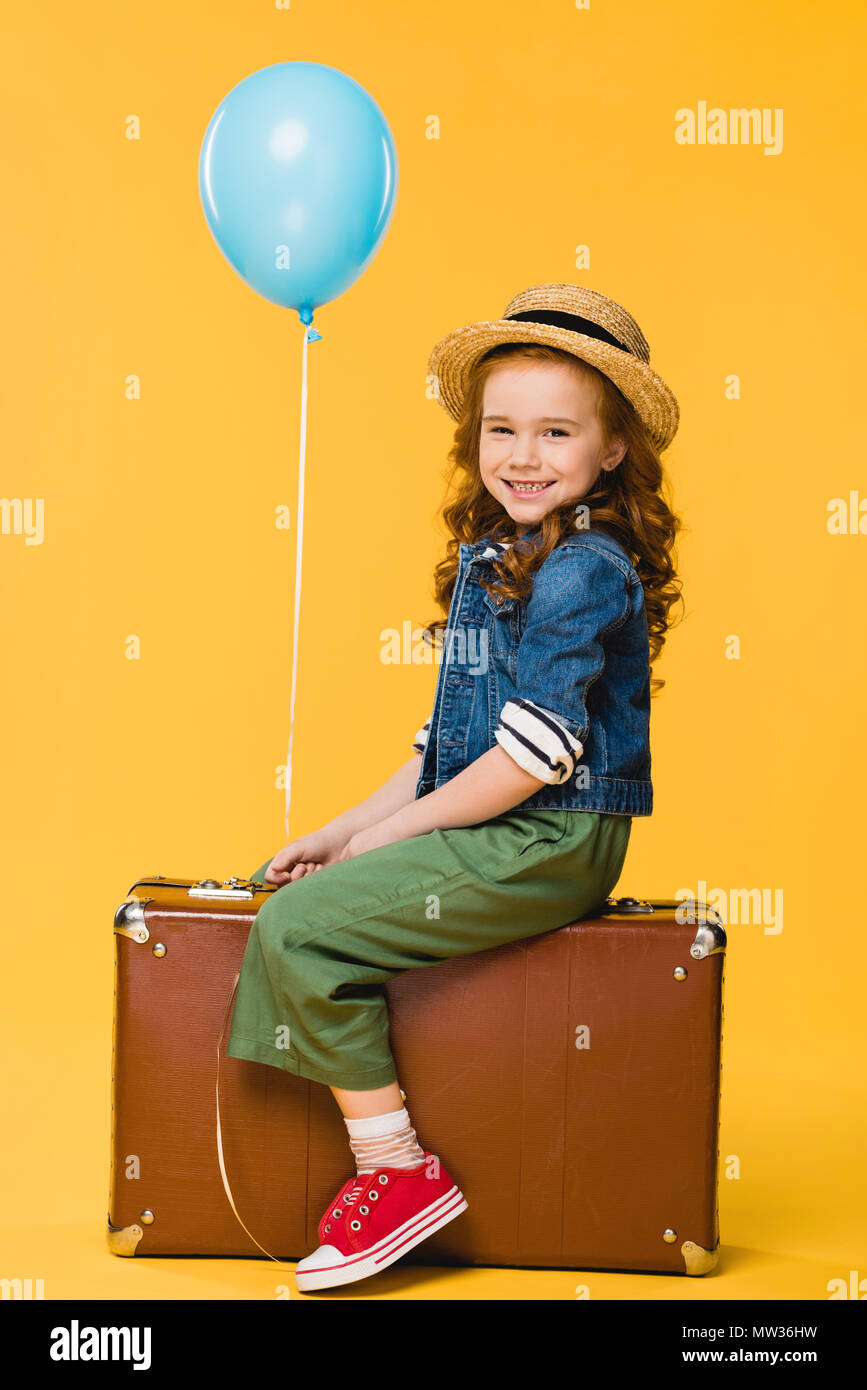 side view of kid with balloon sitting in suitcase isolated on yellow ...