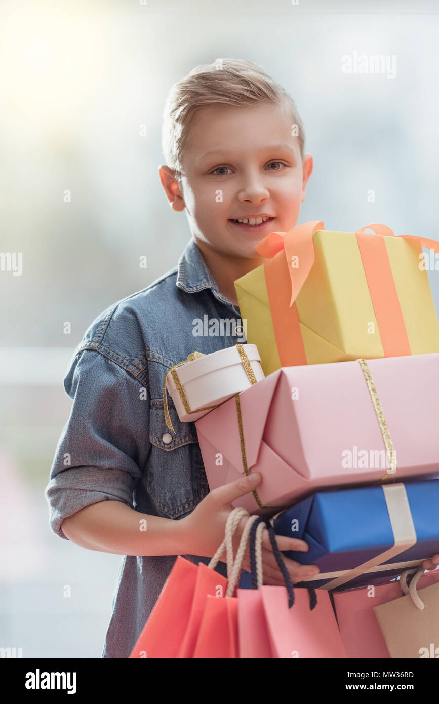 smiling boy holding boxes with colored paper bags in hands at shop ...
