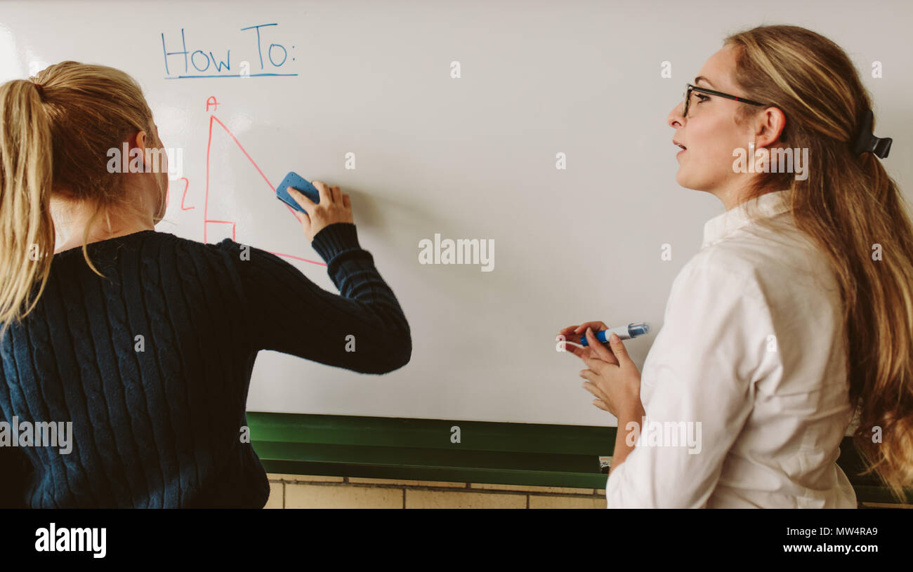 Rear view of young student writing on board with teacher standing by in ...