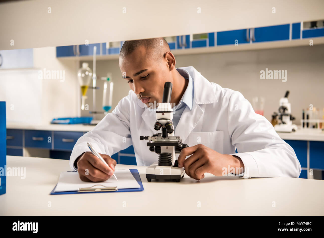 Young technician in lab coat taking notes while doing a sample analysis ...
