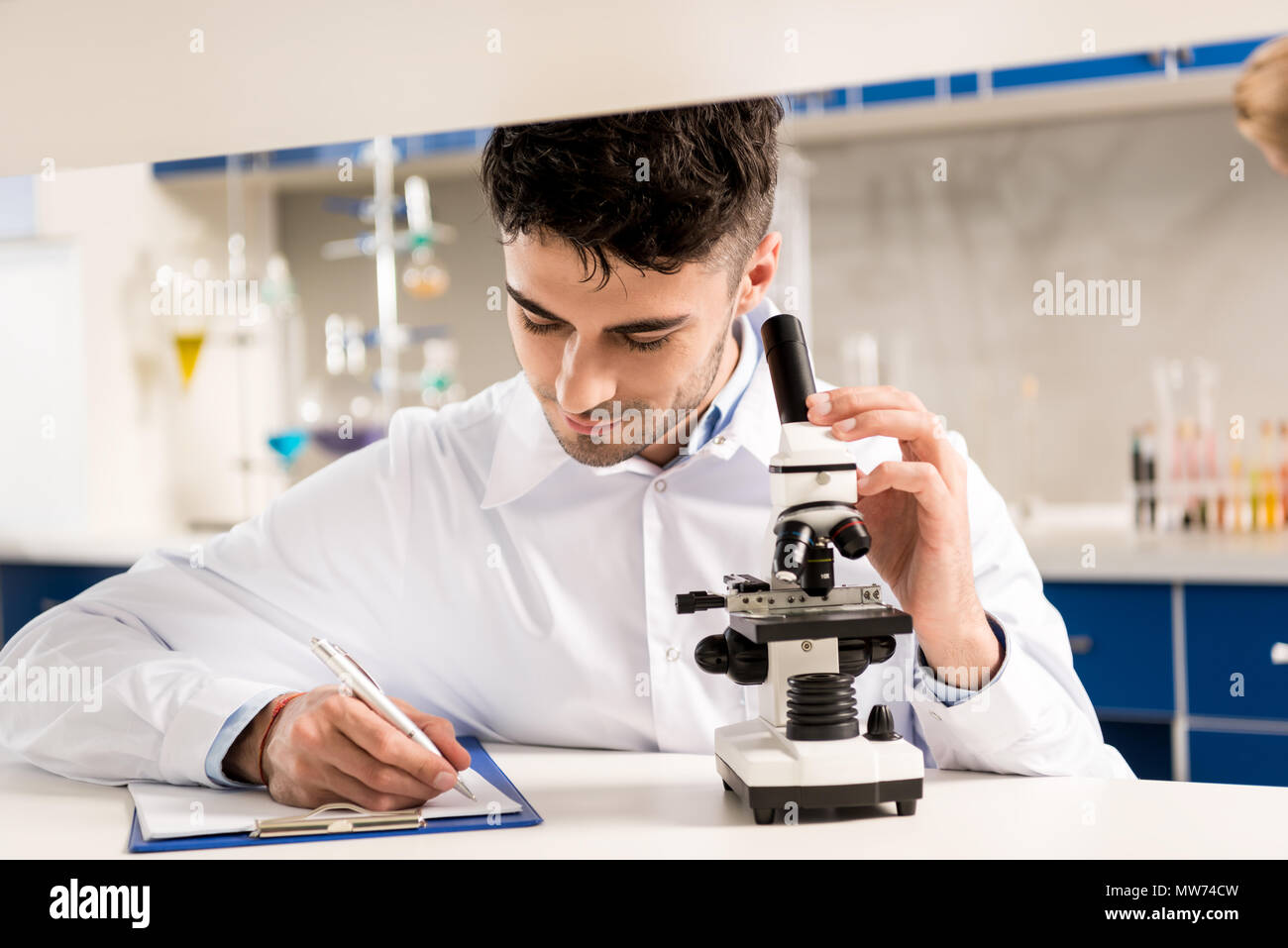 Young lab technician in white coat taking notes and using microscope in ...