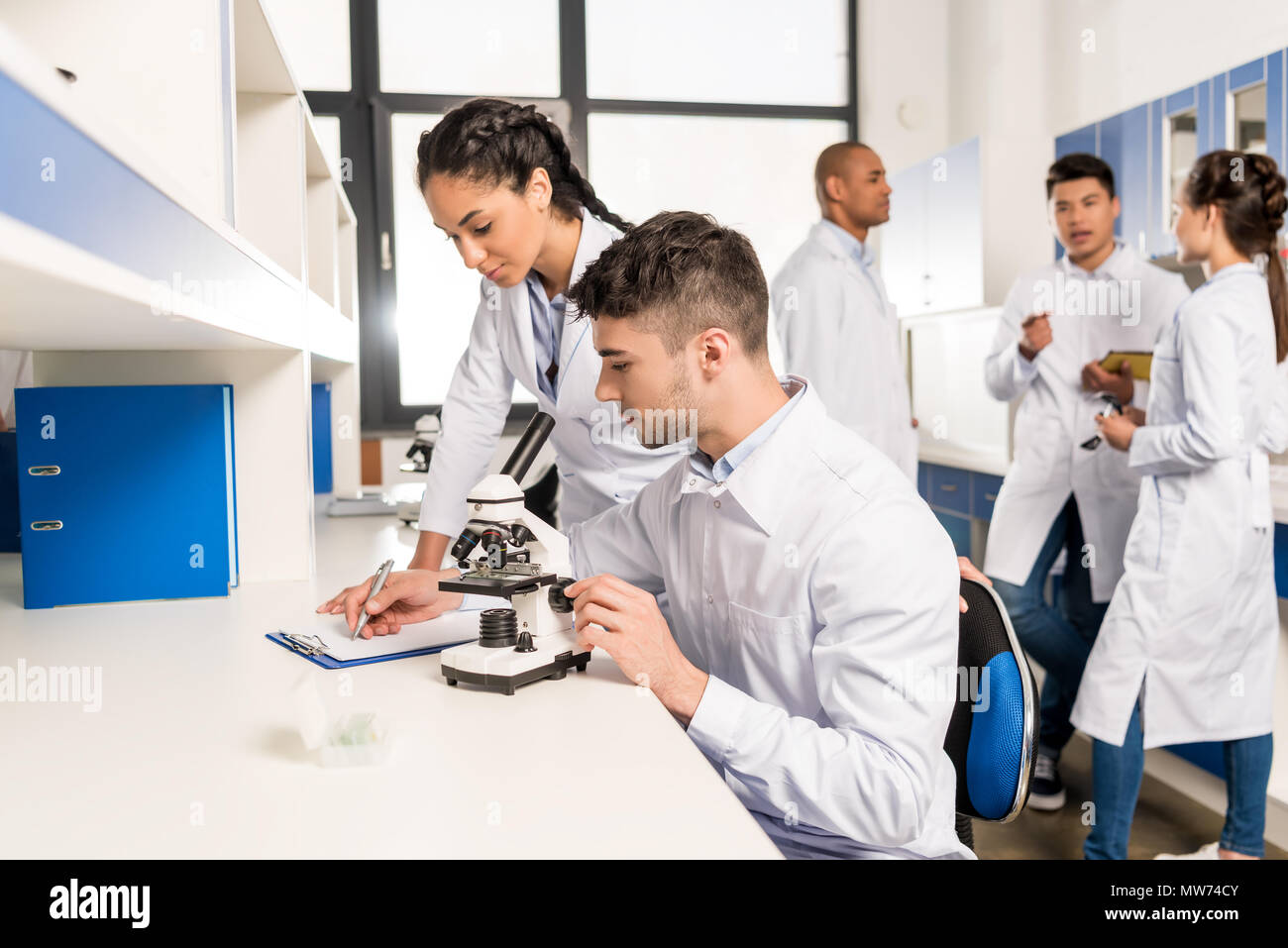 Young lab technicians working with microscope and taking notes on ...
