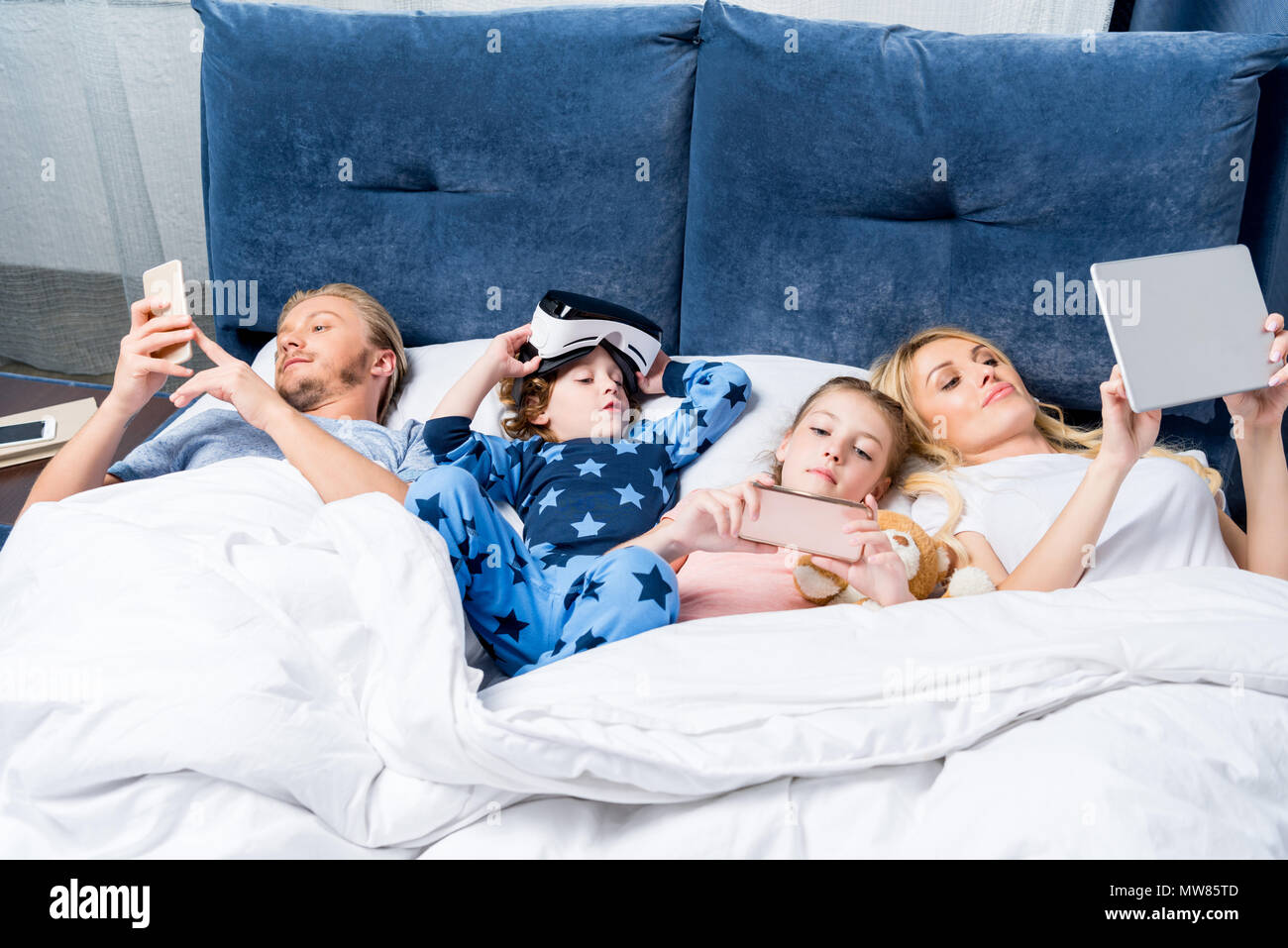 family with two children using digital devices while lying together in ...