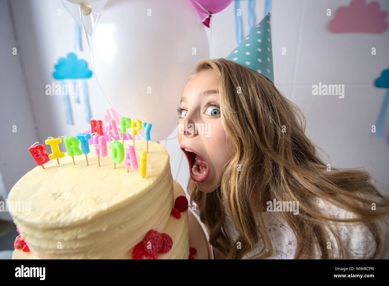 cute little girl in party cap biting birthday cake and looking at ...