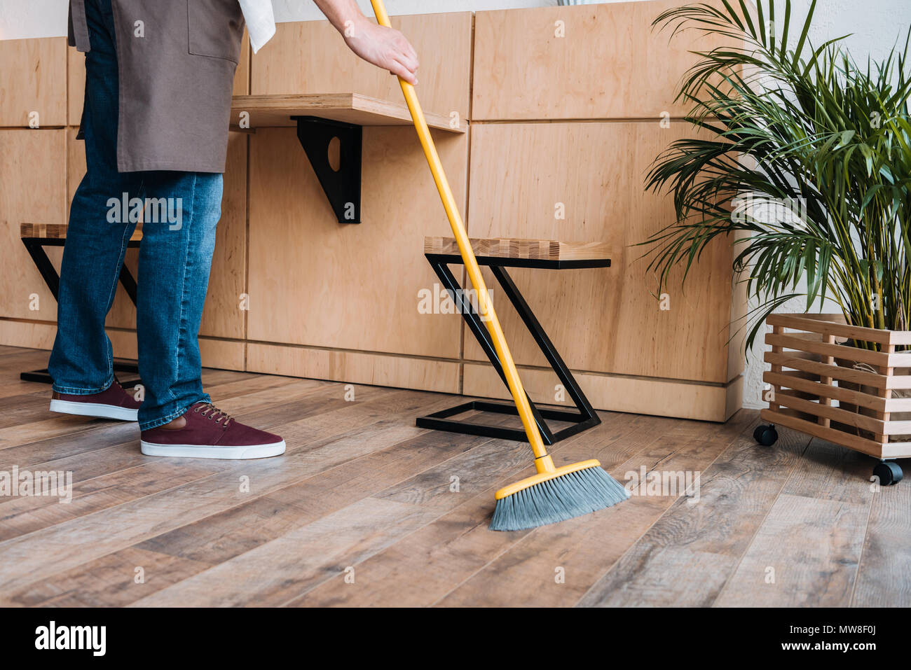 partial view of barista in apron cleaning floor with broom in cafe ...