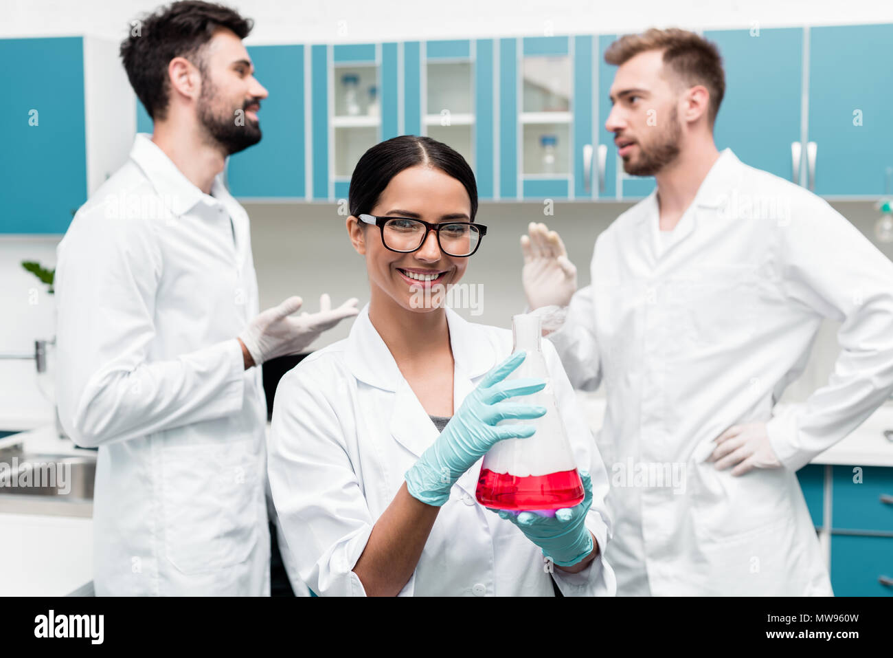 Smiling young chemist holding flask with reagent while colleagues ...