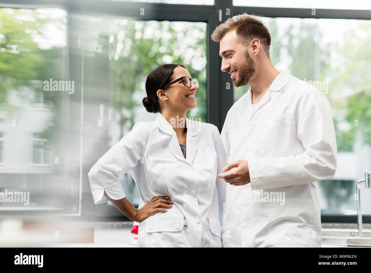 Cheerful young scientists in white coats smiling each other in lab ...