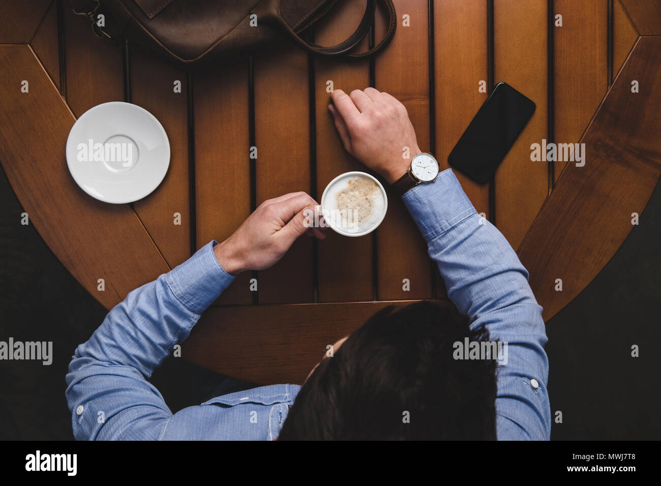 overhead view of man drinking coffee at wooden table with leather bag ...