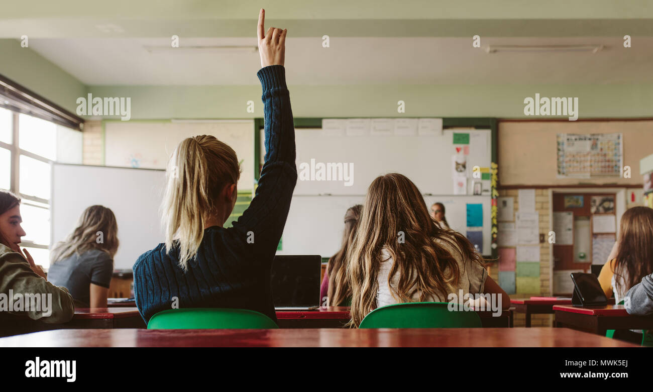 Rear view of female student sitting in the class and raising hand up to ...