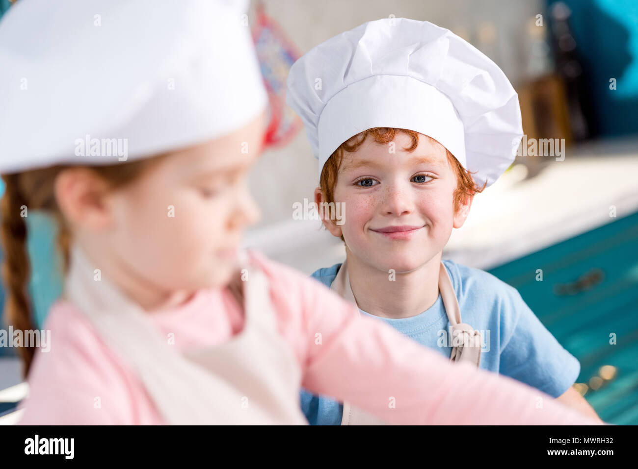 adorable smiling little boy in chef hat looking at friend cooking on ...