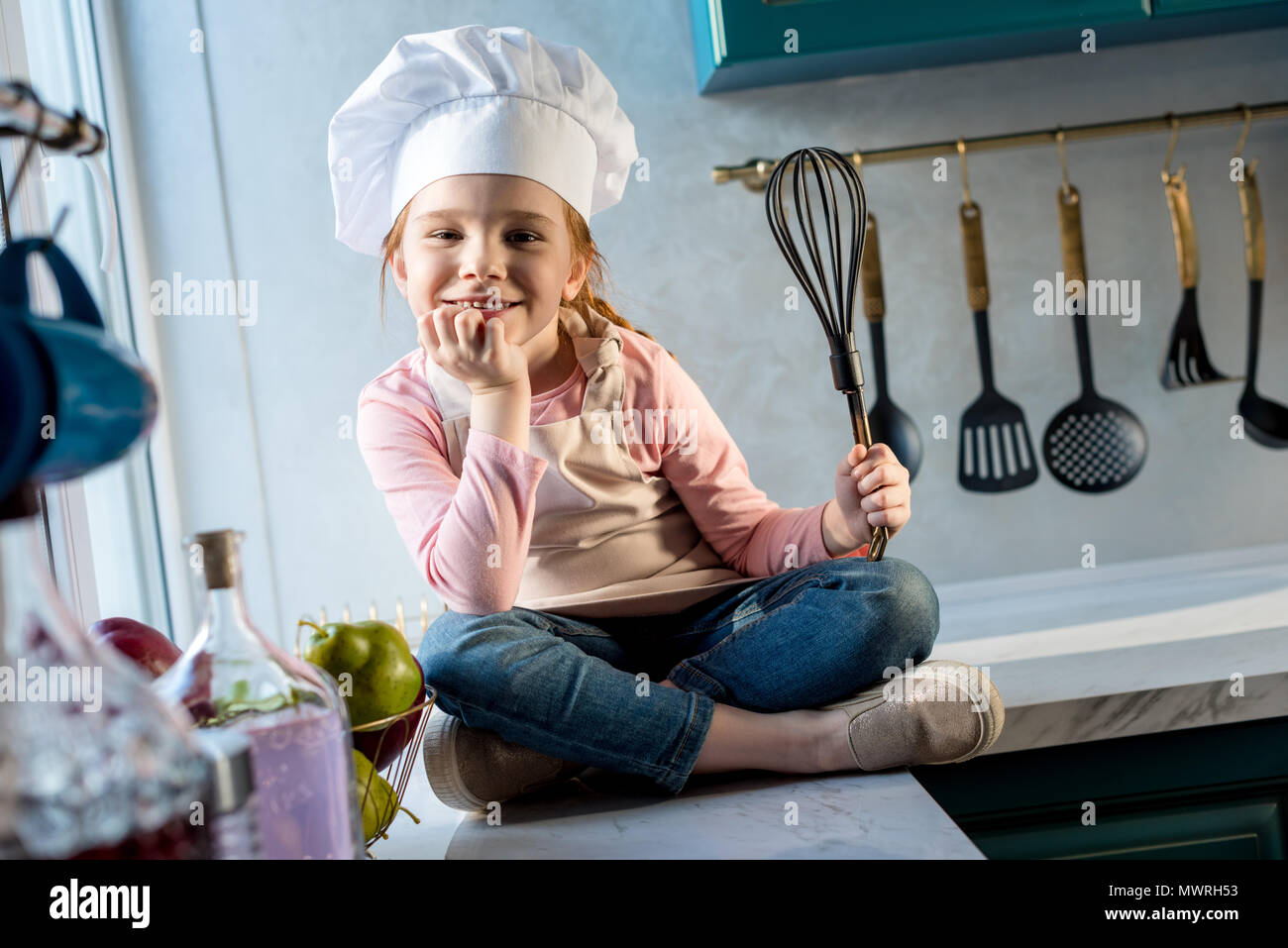 cute child in chef hat holding whisk and smiling at camera in kitchen ...