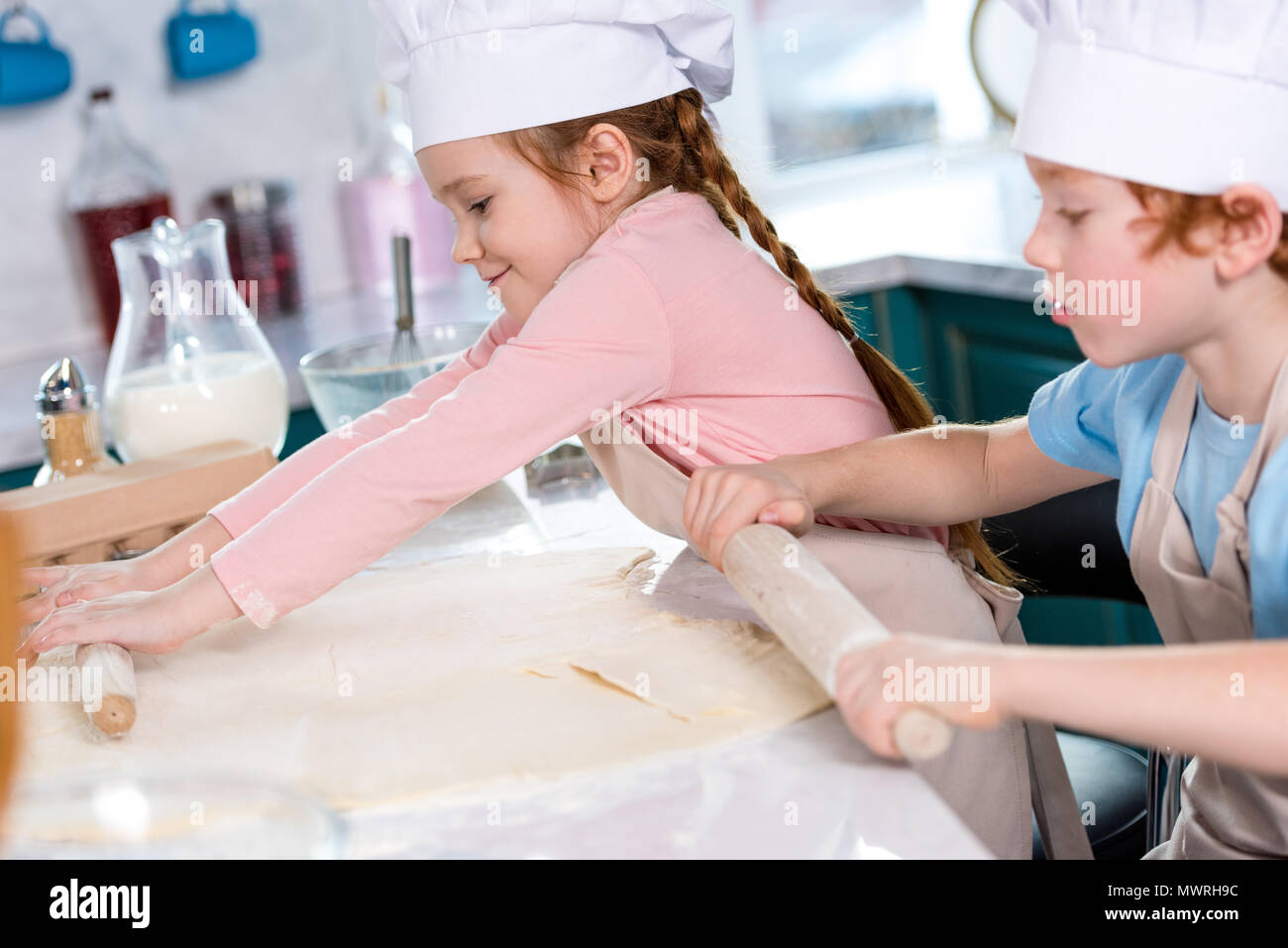 side view of cute little children in chef hats and aprons rolling dough ...