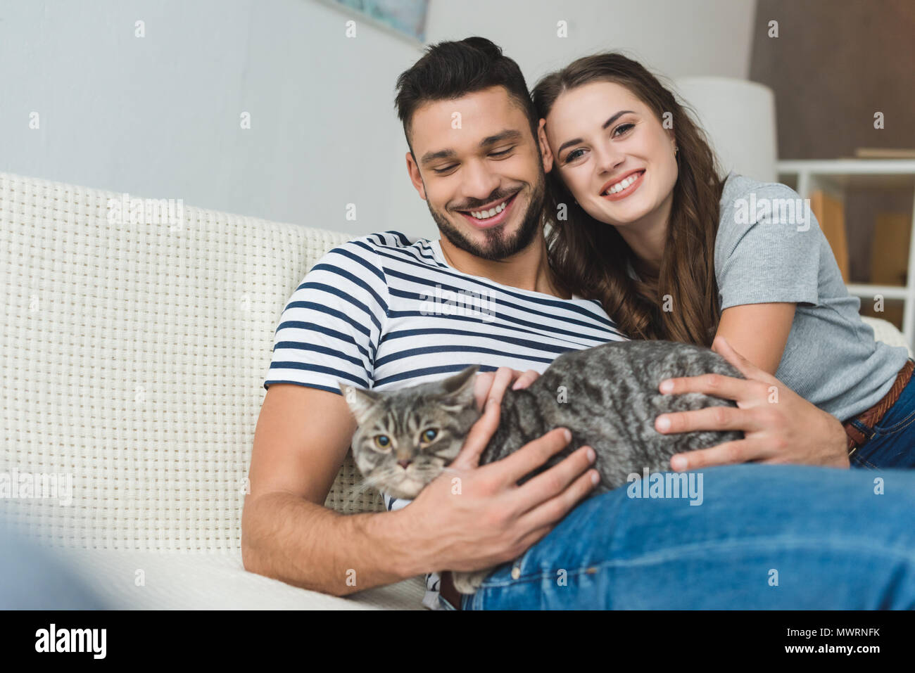 happy young couple with cute tabby cat sitting on couch Stock Photo - Alamy