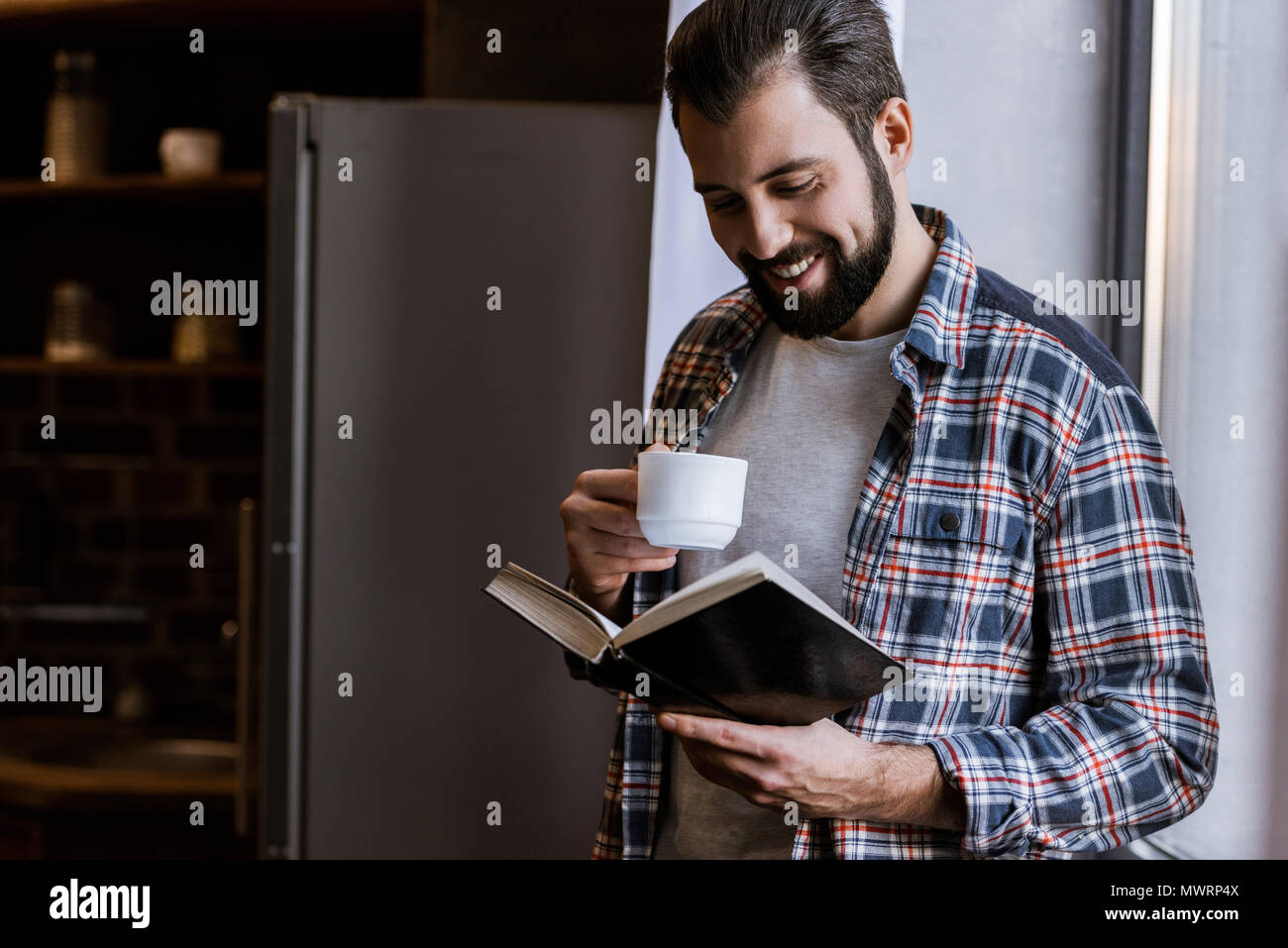 cheerful man with coffee cup standing beside window and reading book ...