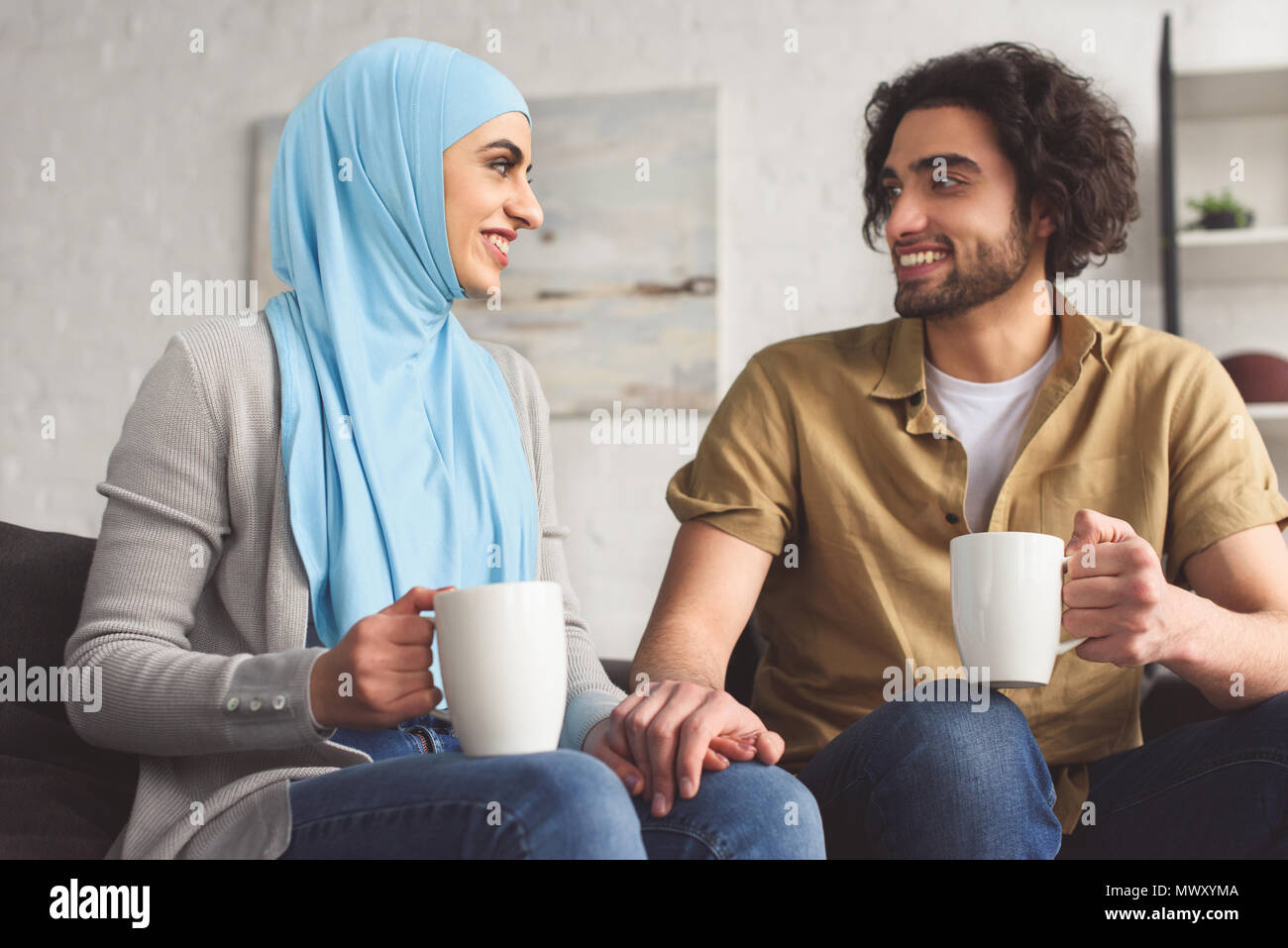 smiling muslim couple holding hands and holding cups of coffee at home ...