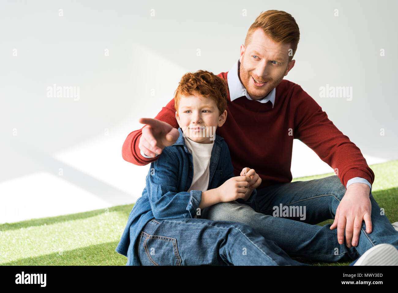 happy redhead father and son sitting together and looking away on grey ...