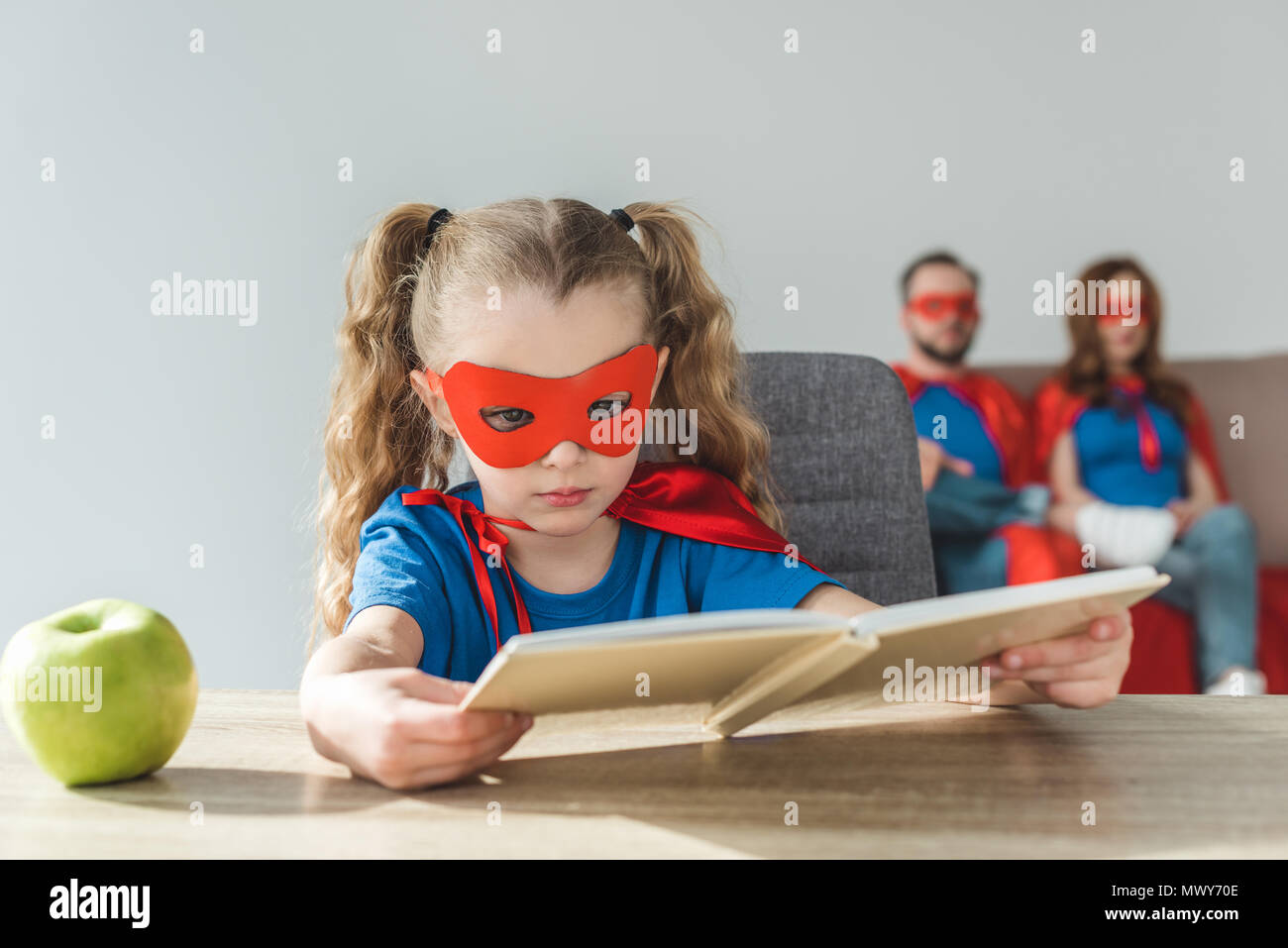 girl in superhero costume reading book while super parents sitting ...