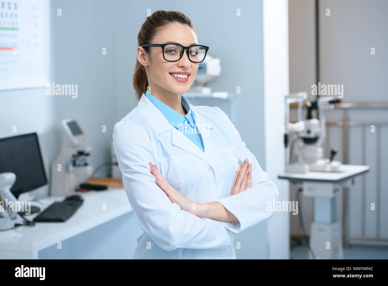 attractive professional ophthalmologist in glasses standing with crossed arms in optical clinic ...