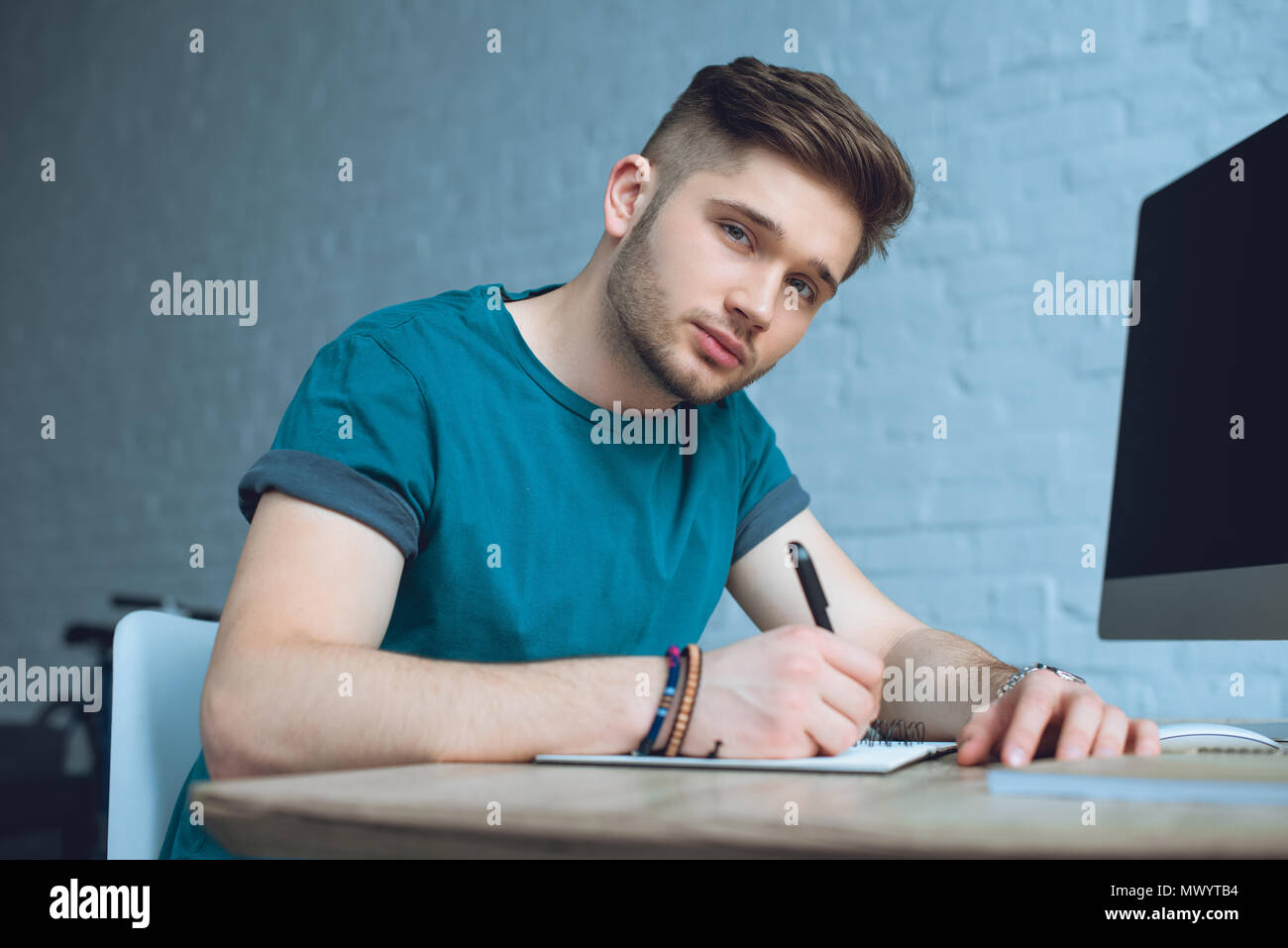 handsome young man taking notes and looking at camera while working at ...