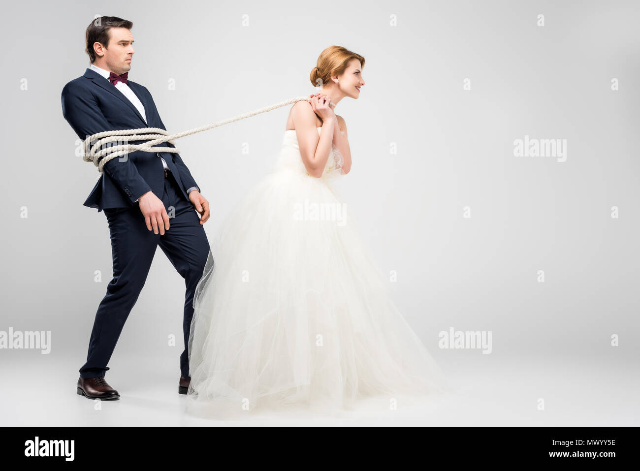 bride in wedding dress pulling groom bound with rope, isolated on grey ...