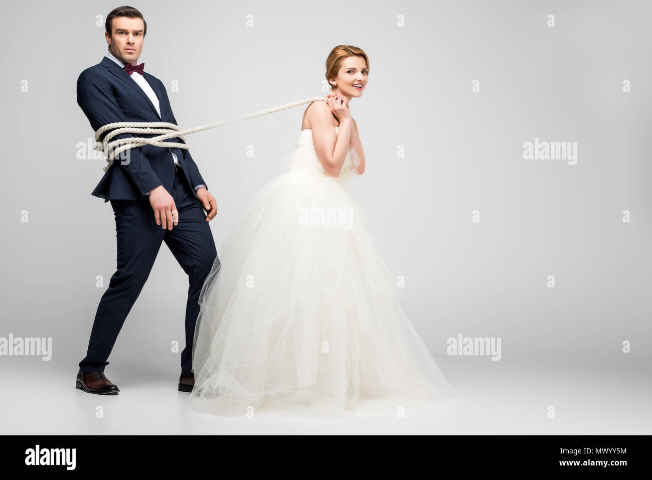beautiful bride pulling groom bound with rope, isolated on grey ...