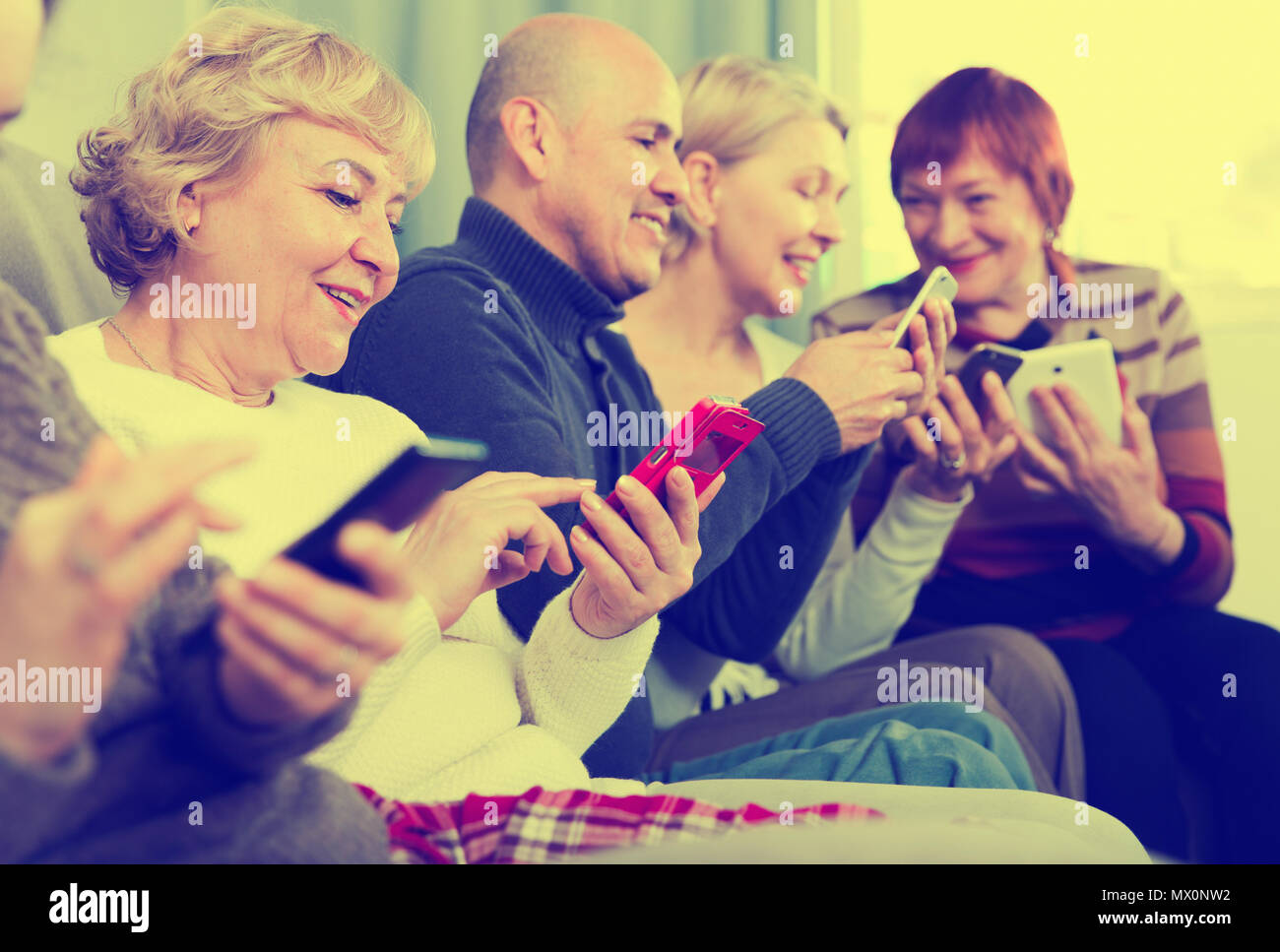 Mature friends are resting together and looking at their phones at home ...