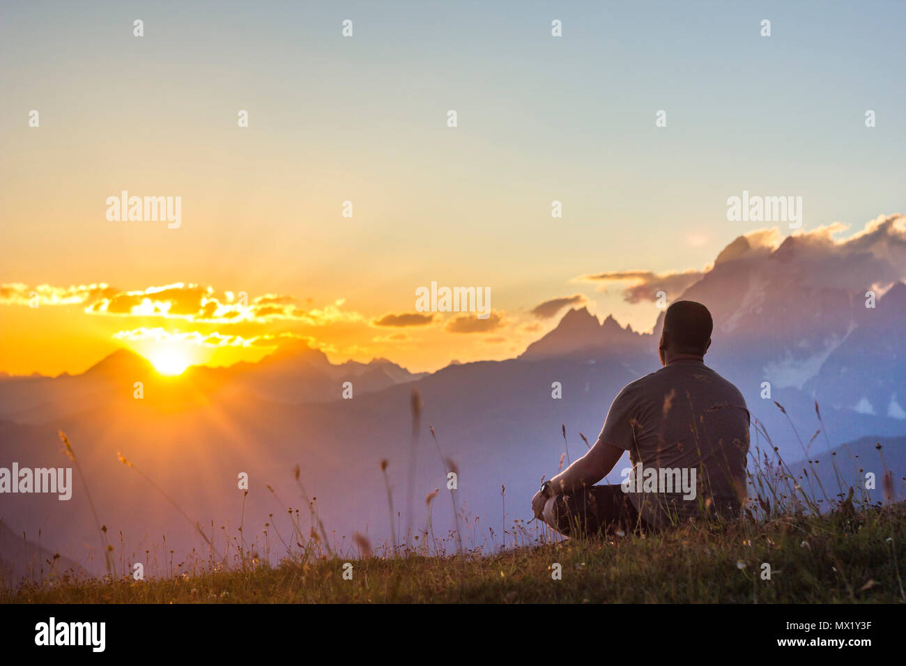 man sitting on grass at sunset in mountains thinking and looking on sun ...