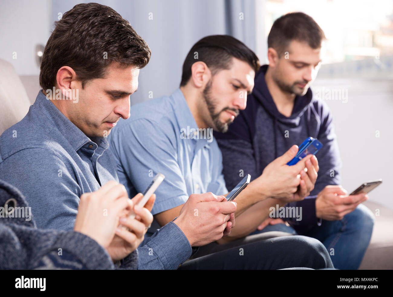 Positive men sitting on couch at home absorbedly looking at phones ...
