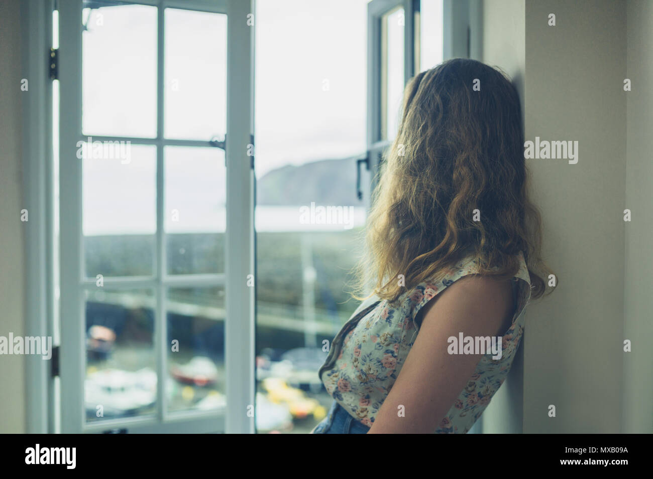 A young woman is sitting and resting by the window of a country house ...