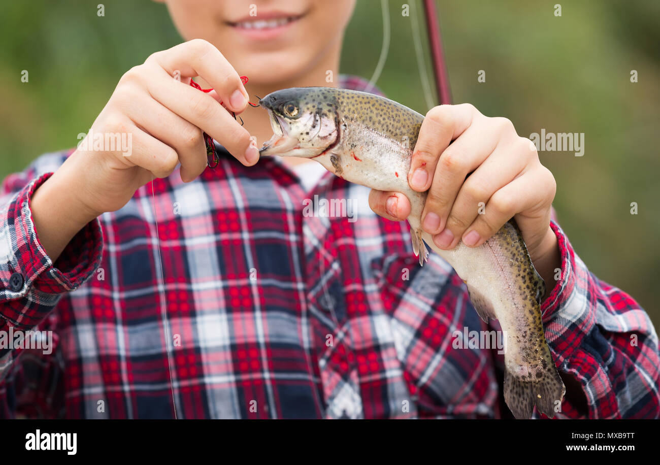 portrait of smiling teenager boy holding catch fish on hook on lake ...