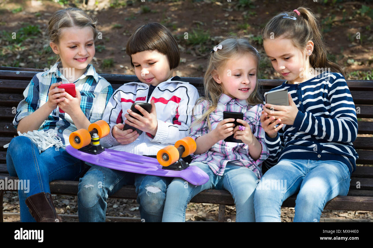 Happy smiling children sitting on bench and playing with mobile devices ...