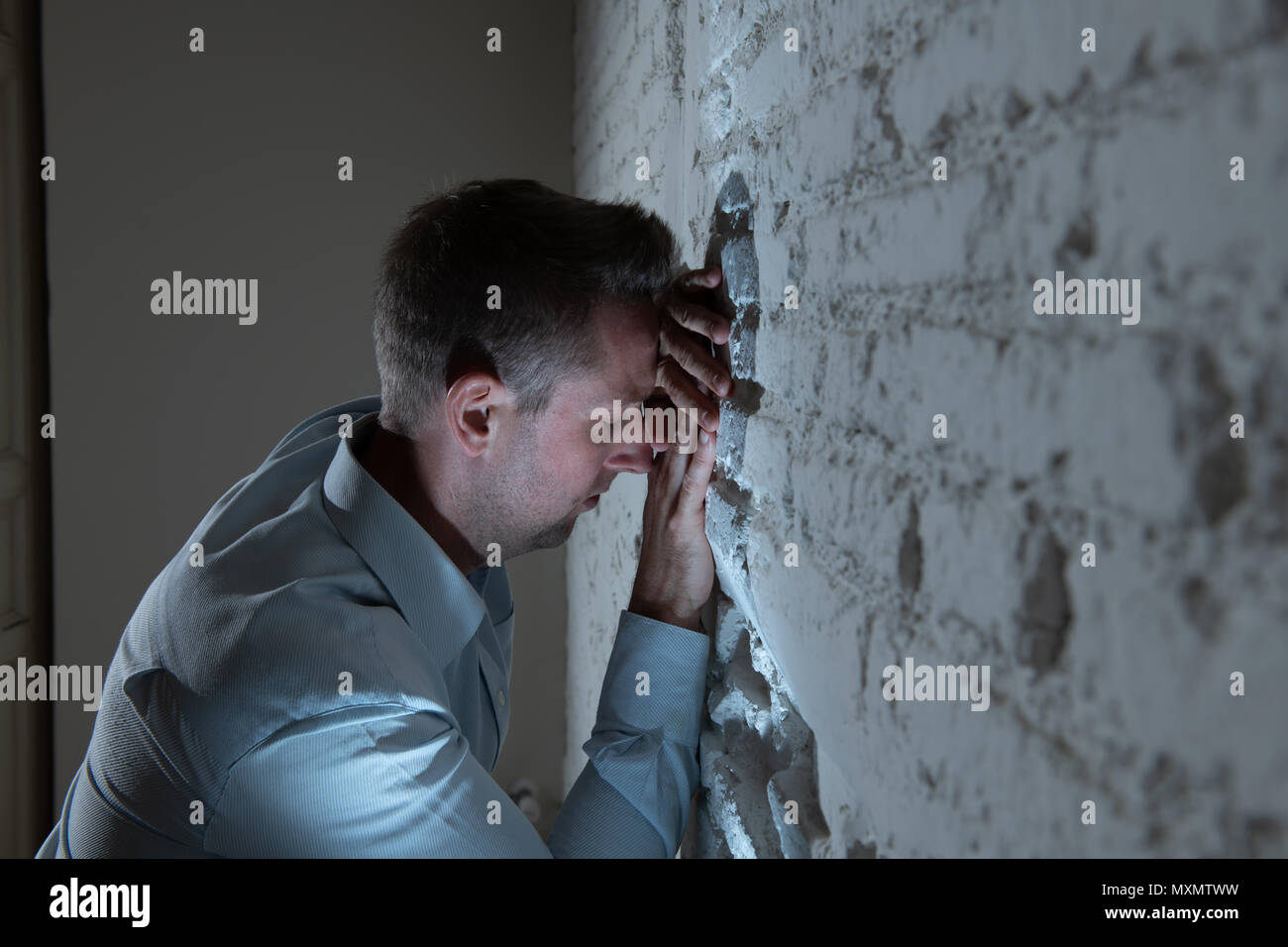 depressed man leaning up on a white wall in dark room at home, feeling ...