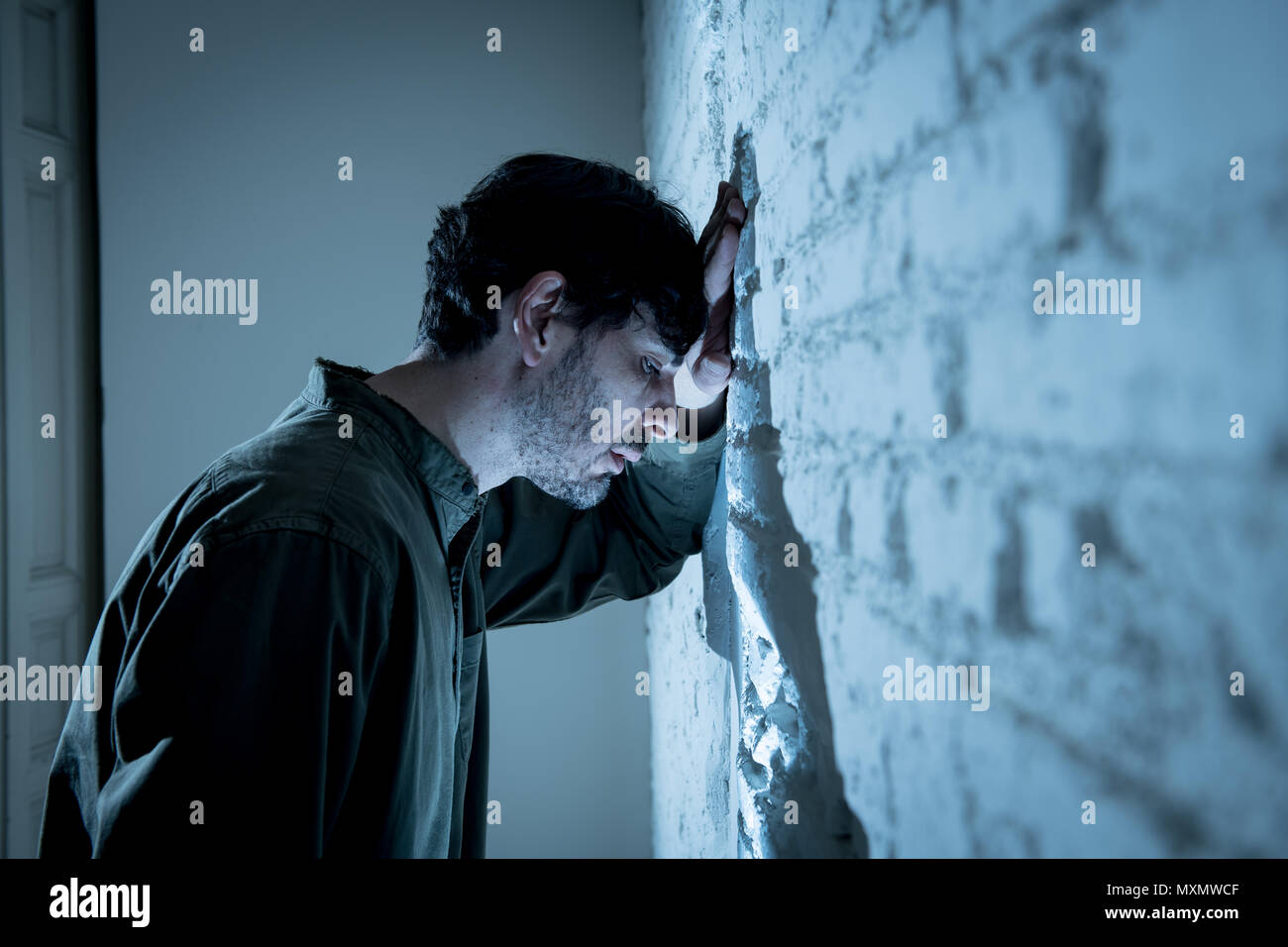 depressed man leaning up on a white wall in dark room at home, feeling ...