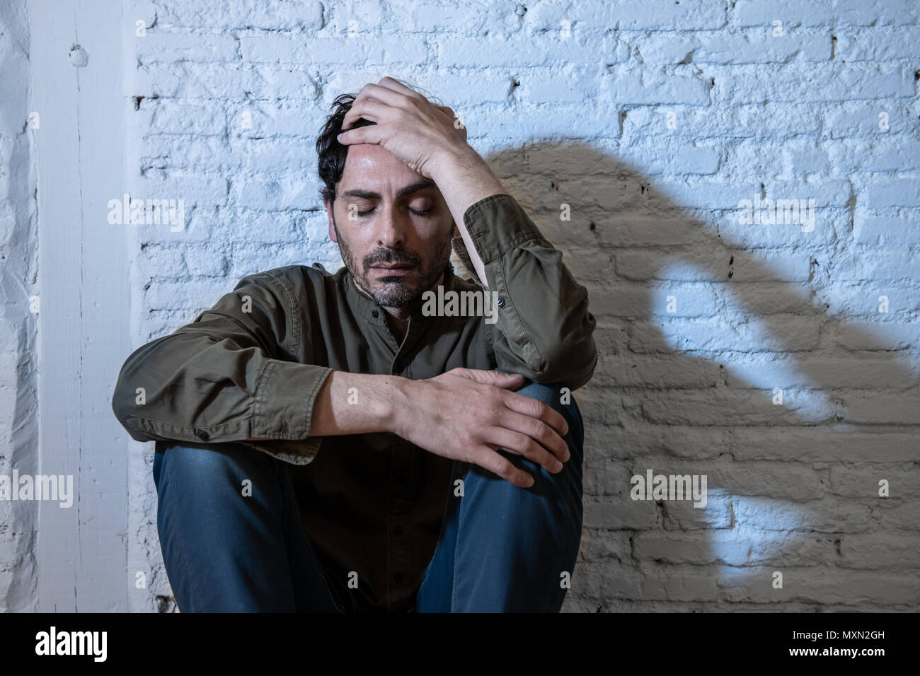 young depressed man sitting against a white wall at home with a shadow ...