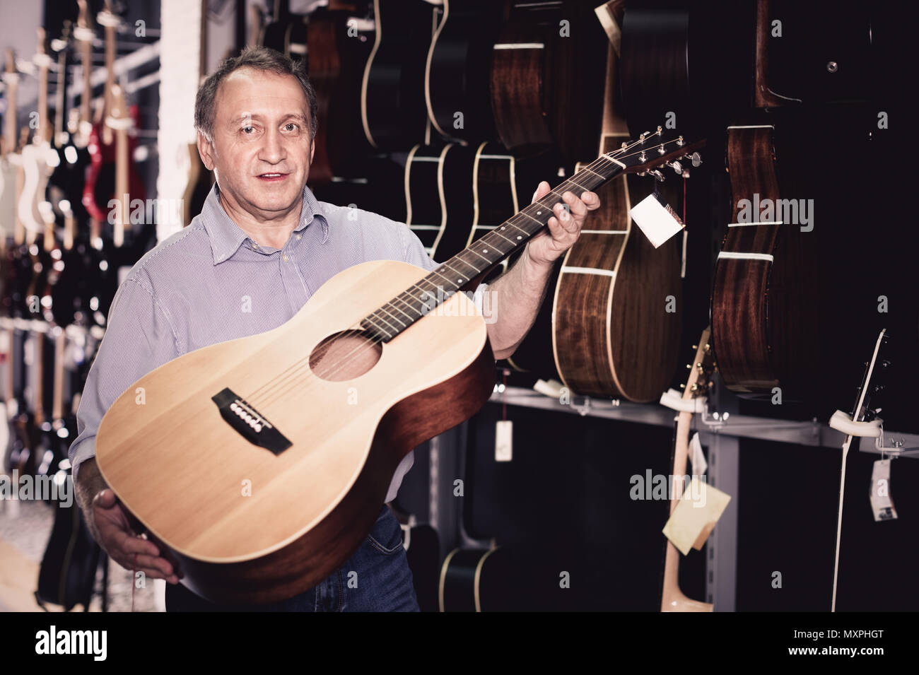 Adult smiling guitarist is standing with acoustic guitar in music store ...