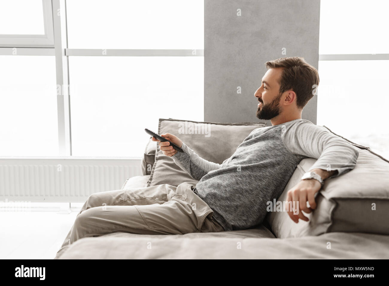 Portrait of a cheerful young man holding TV remote control while ...