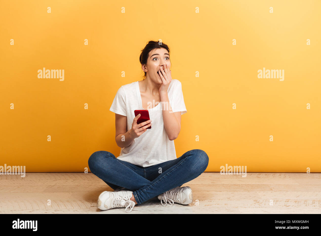 Portrait of a shocked young woman holding mobile phone while sitting ...