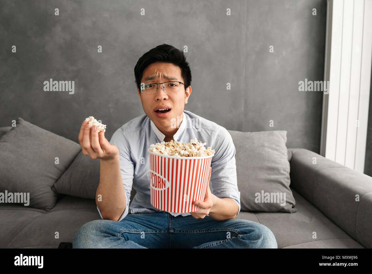 Portrait of a disappointed young asian man eating popcorn while sitting ...