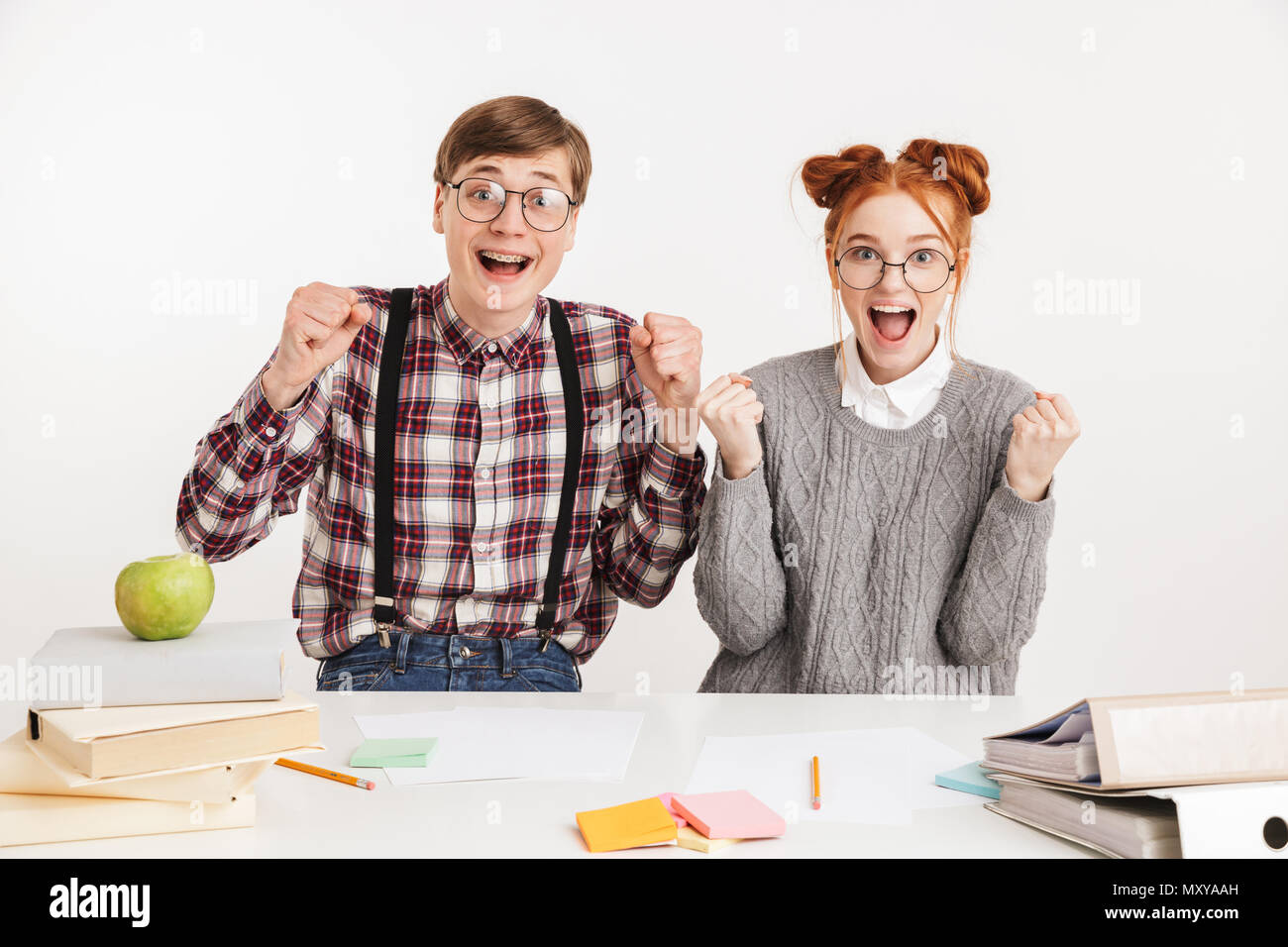 Funny couple of school nerds celebrating while sitting at the desk isolated over white ...