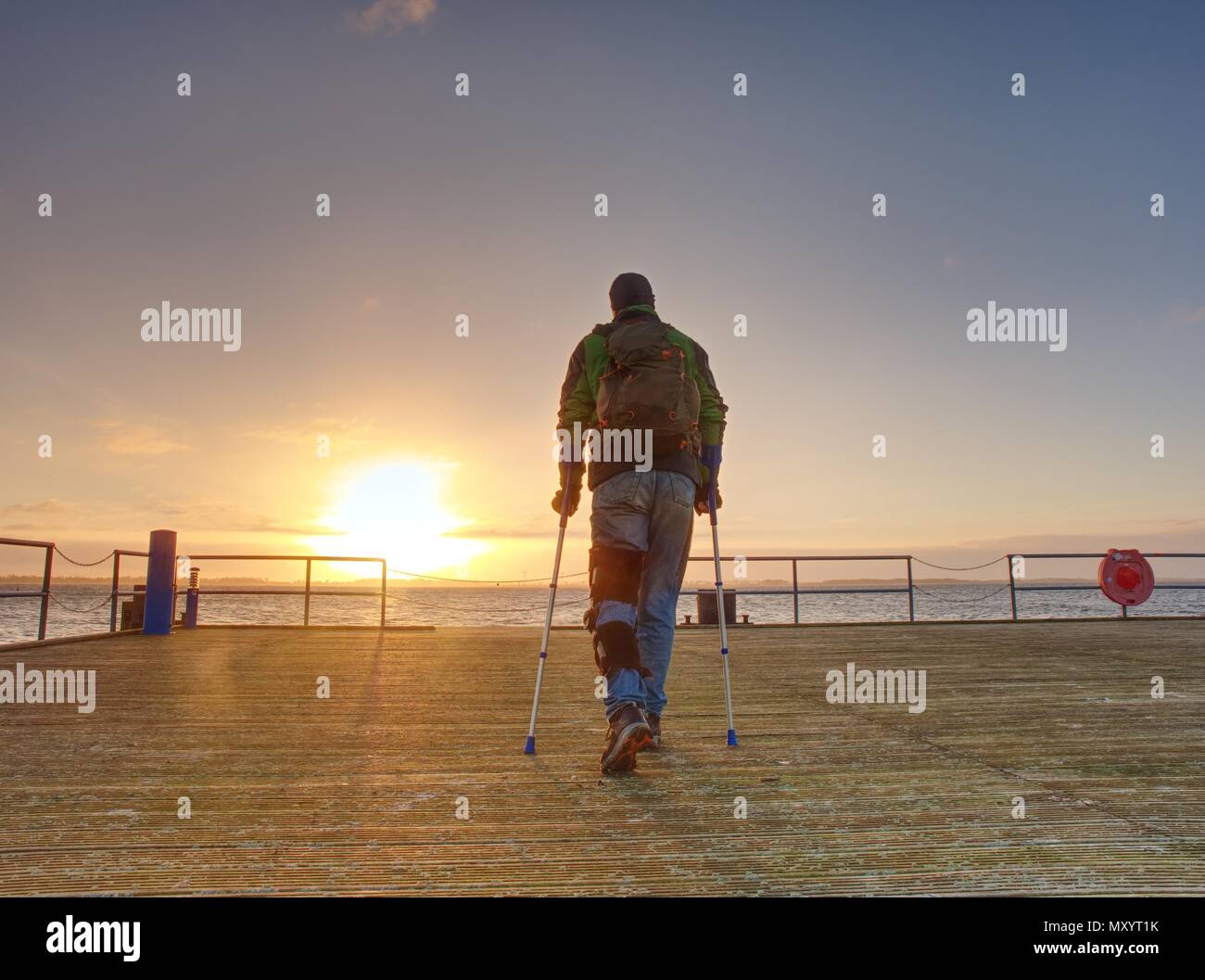 Hurt man with hooded jacket and forearm crutches looking sadly into sea ...