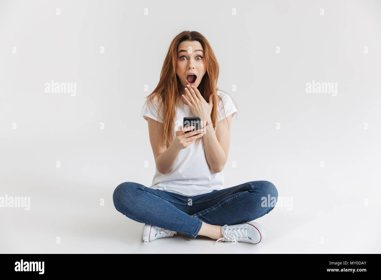 Portrait of a shocked young girl using mobile phone while sitting with ...