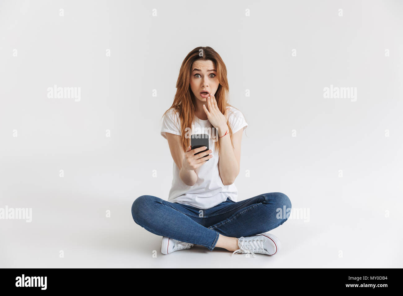Portrait of a shocked young girl using mobile phone while sitting with ...