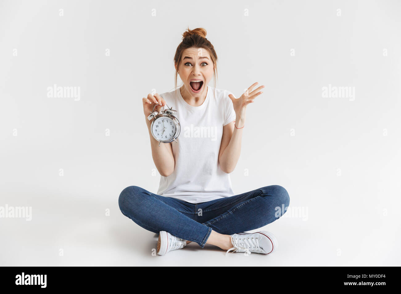 Portrait of a shocked young girl holding alarm clock while sitting with ...