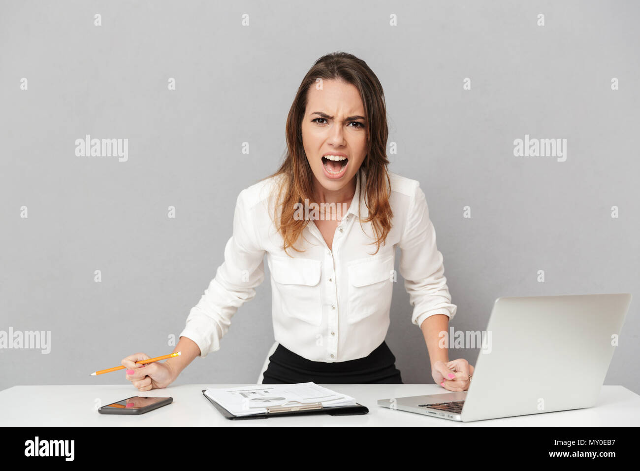 Portrait of a furious young business woman sitting at the office desk ...