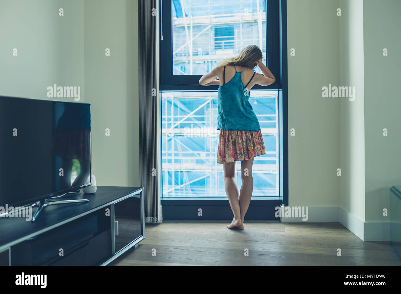 A young woman is standing by the window in her high rise apartment in ...